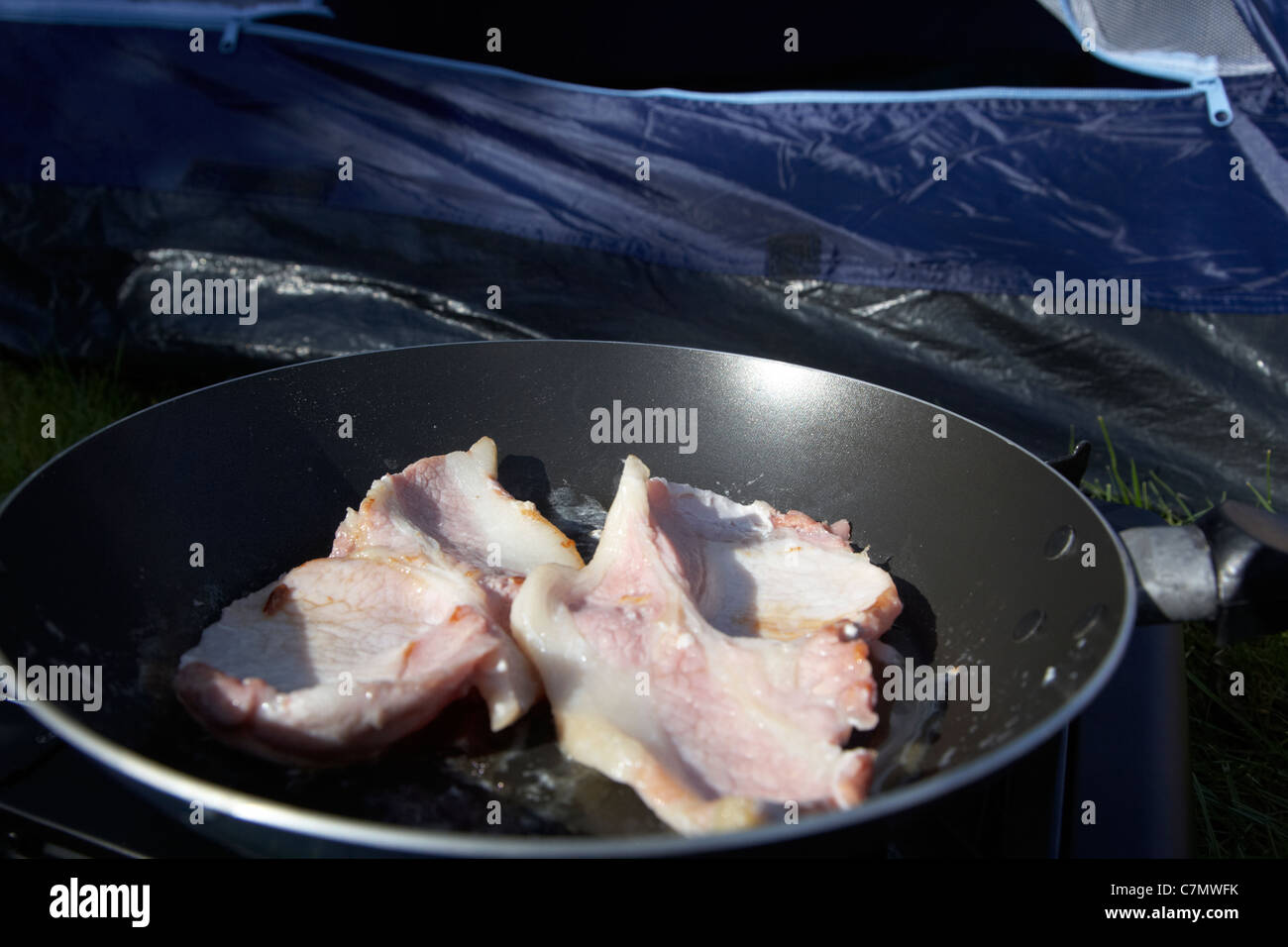 frying bacon on a small portable gas cooker in front of an open tent ...