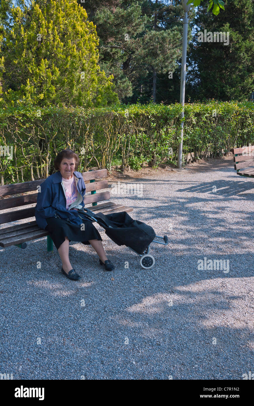 An elderly female senior citizen sits and rests on a park bench to rest ...