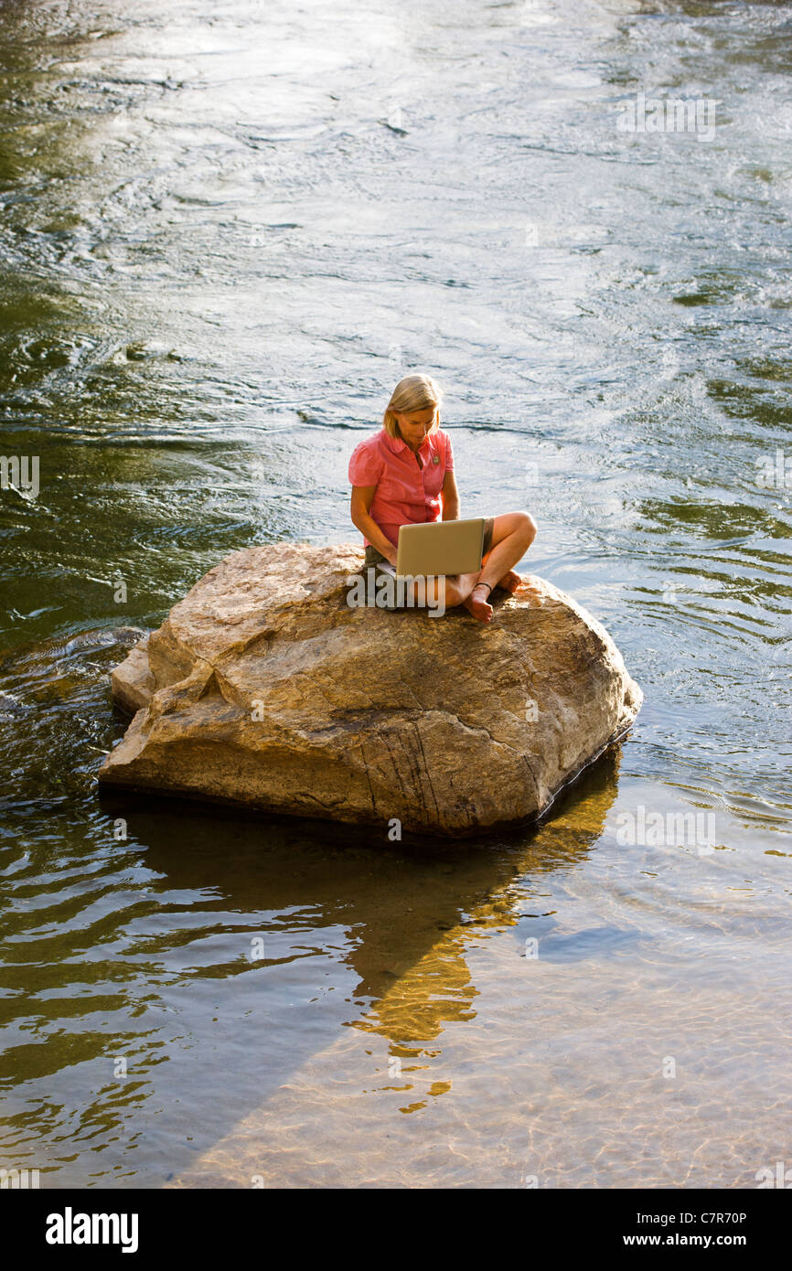 Lone woman sitting on a rock in a river working on a laptop computer ...