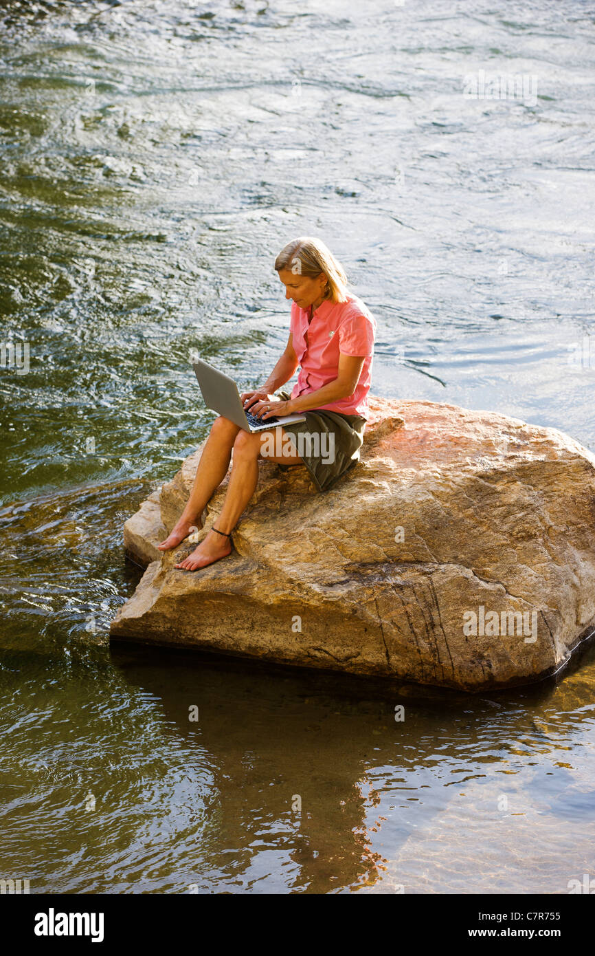 Lone woman sitting on a rock in a river working on a laptop computer ...