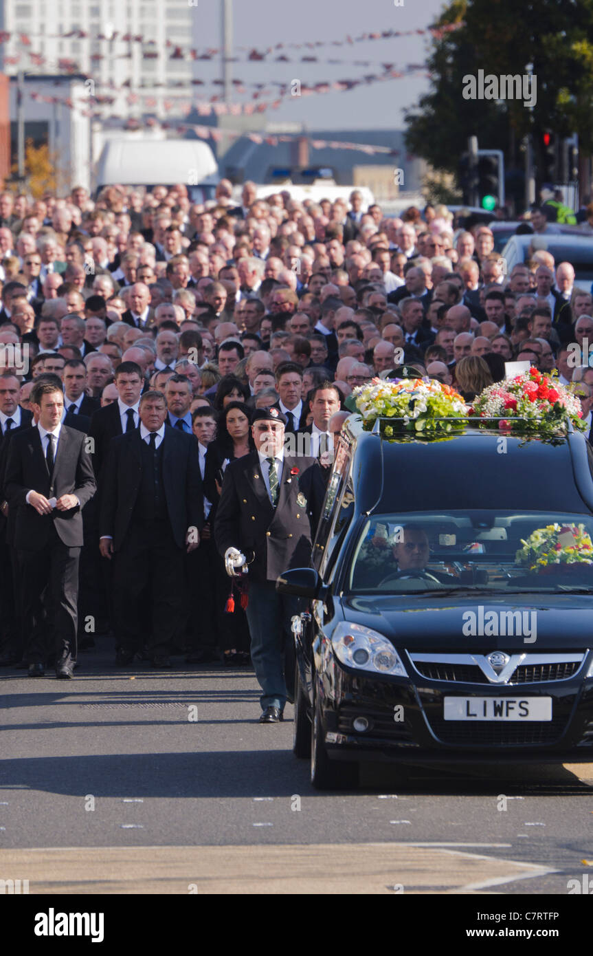 A very large crowd walking behind a hearse on a street during a funeral ...