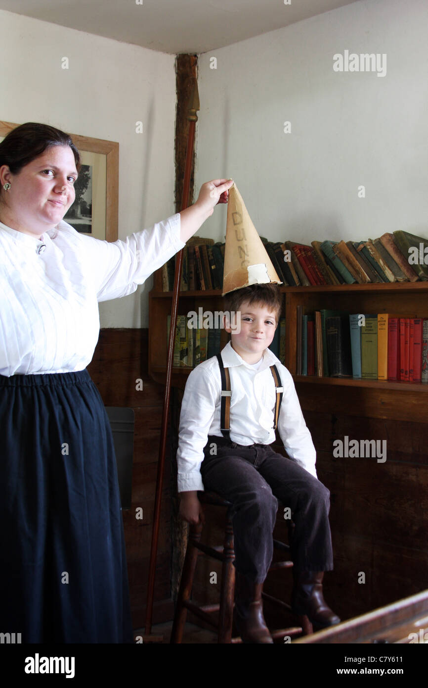 A teacher holding a DUNCE hat on a male child in a historic schoolroom ...