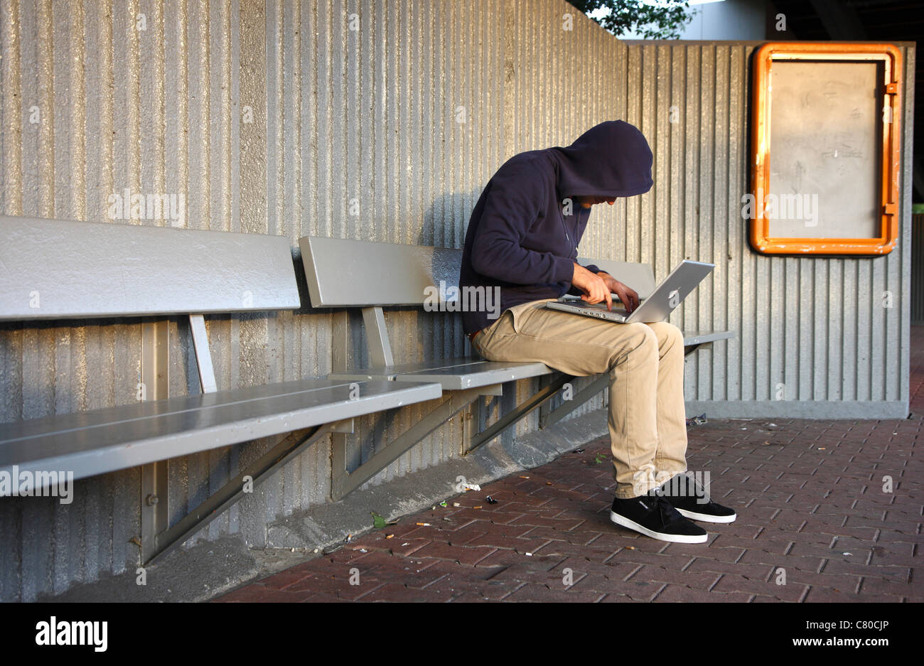 Computer user, hacker, sits conspiratorially, outside, in a bus shelter ...