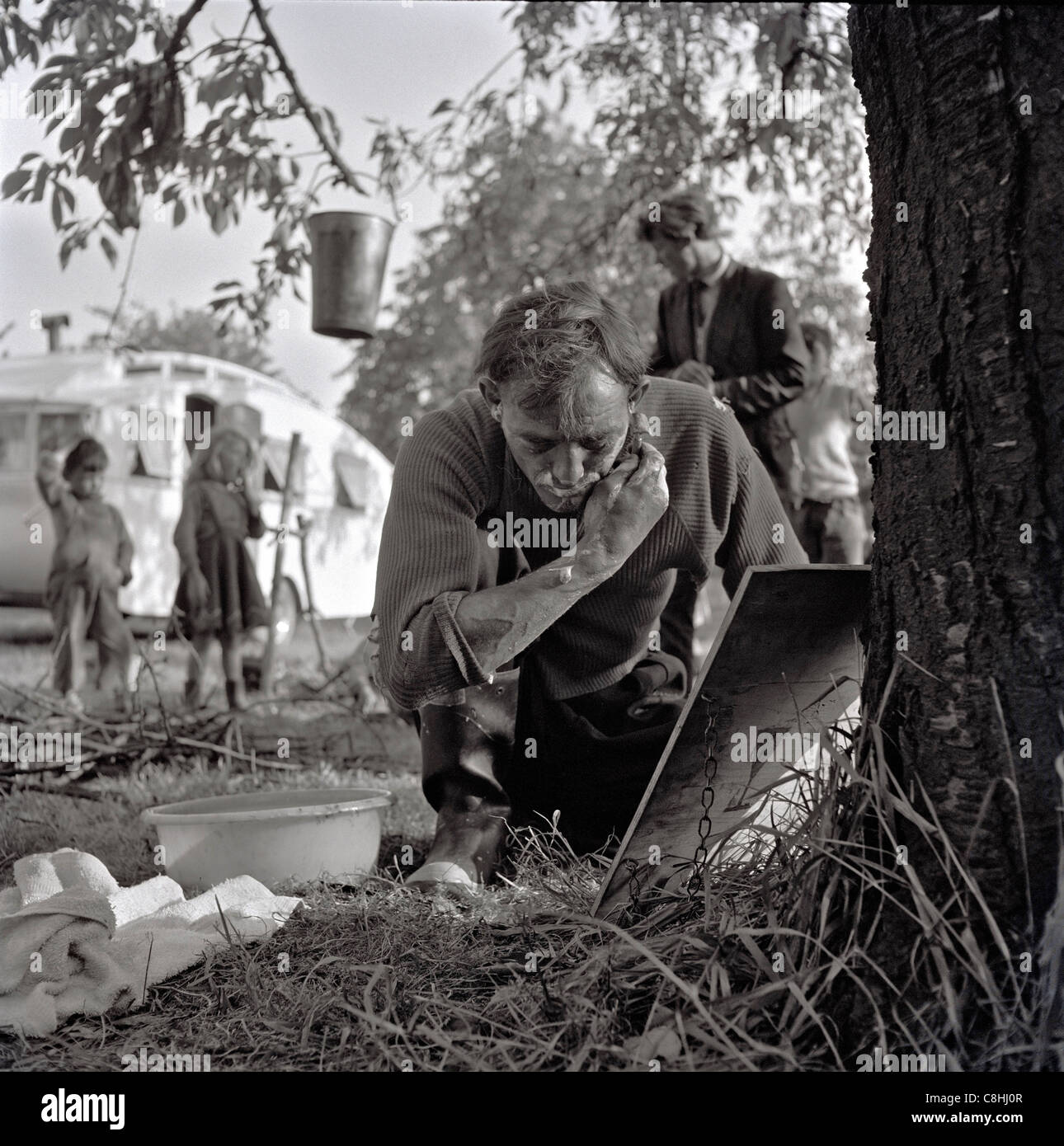 Young gypsy man shaving outdoors with mirror leaning against a tree ...