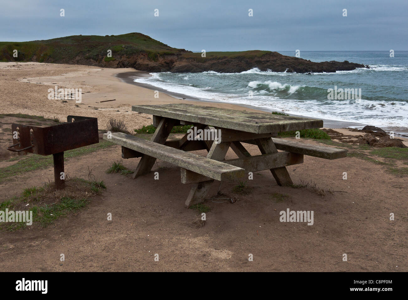 A rustic picnic table and barbecue with a view of ocean waves at a ...