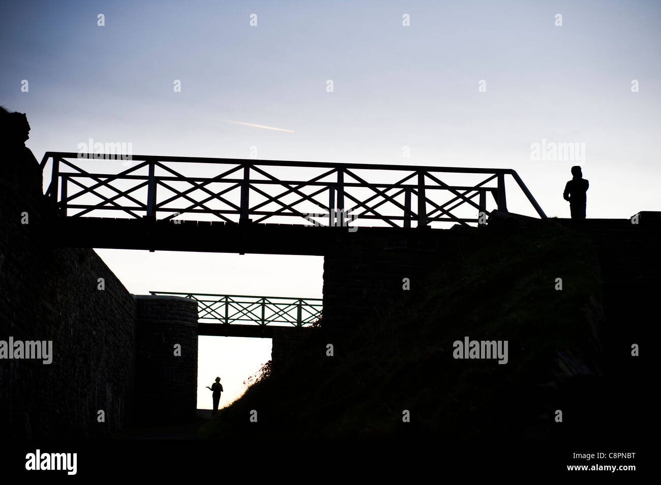 Two separate people standing, and footbridges, silhouetted at dusk, UK ...