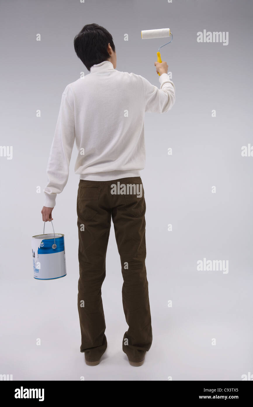 Portrait of young man holding paint roller and paint can, rear view ...