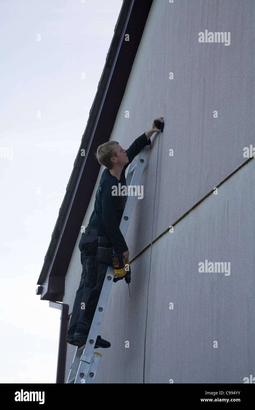 UK Young electrician up a ladder fixing a cable onto wall of domestic ...