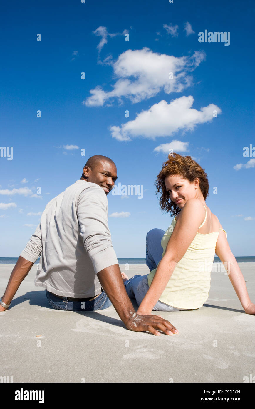 Rear view of a romantic young couple sitting on beach, turning around ...