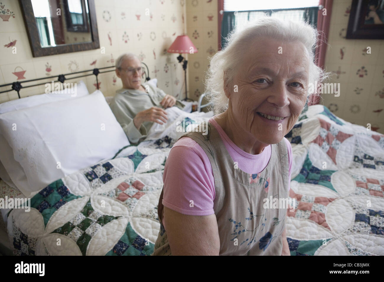 Smiling senior woman sitting on bed with bedridden senior man Stock ...