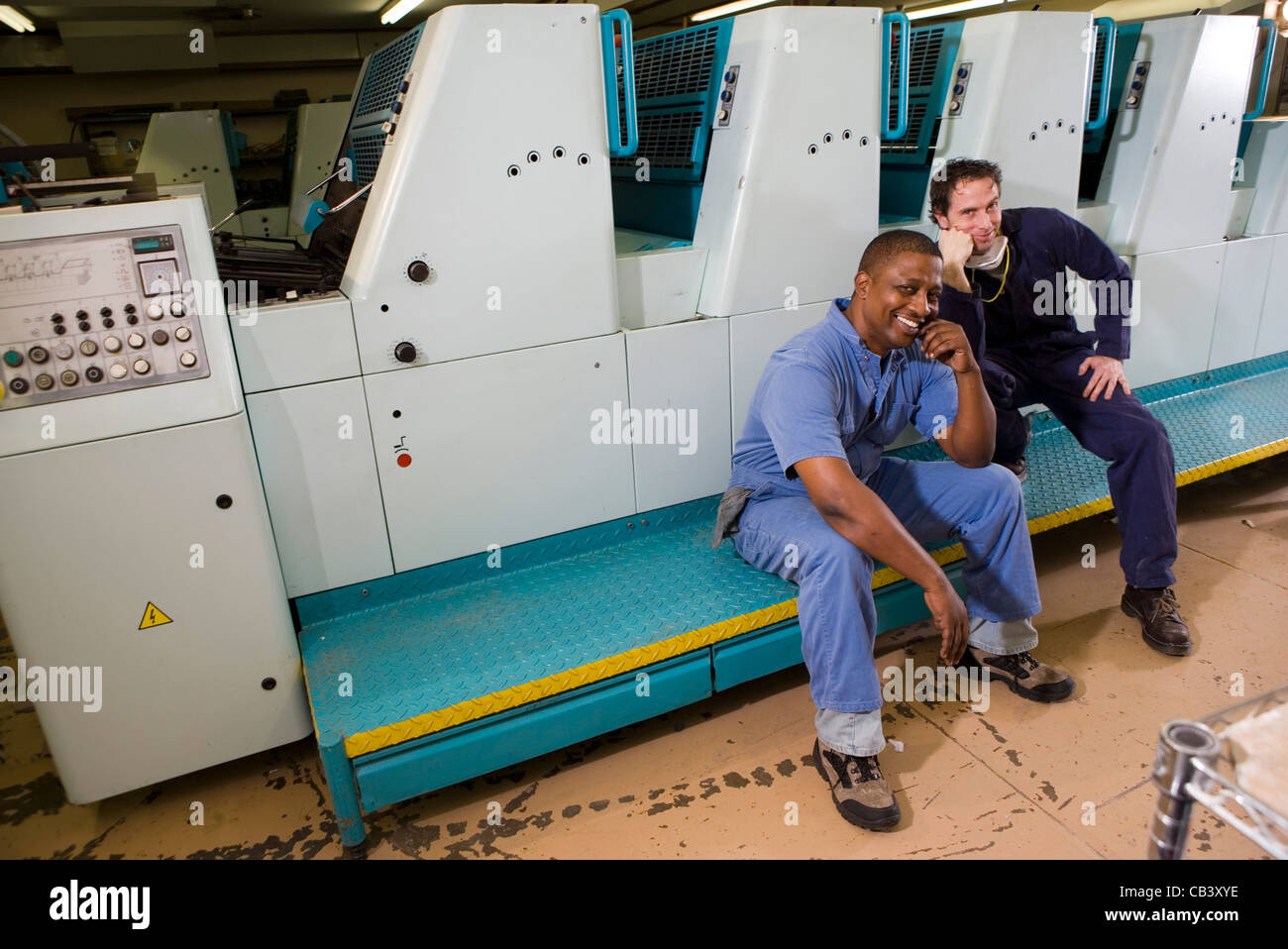 Portrait of two men in coveralls sitting in front of a printing machine ...