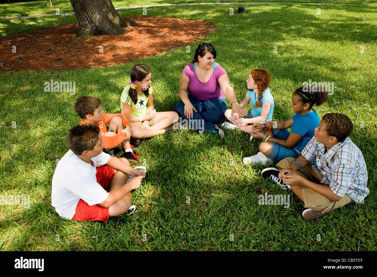 Children sitting in a circle on the grass with their teacher at a park ...