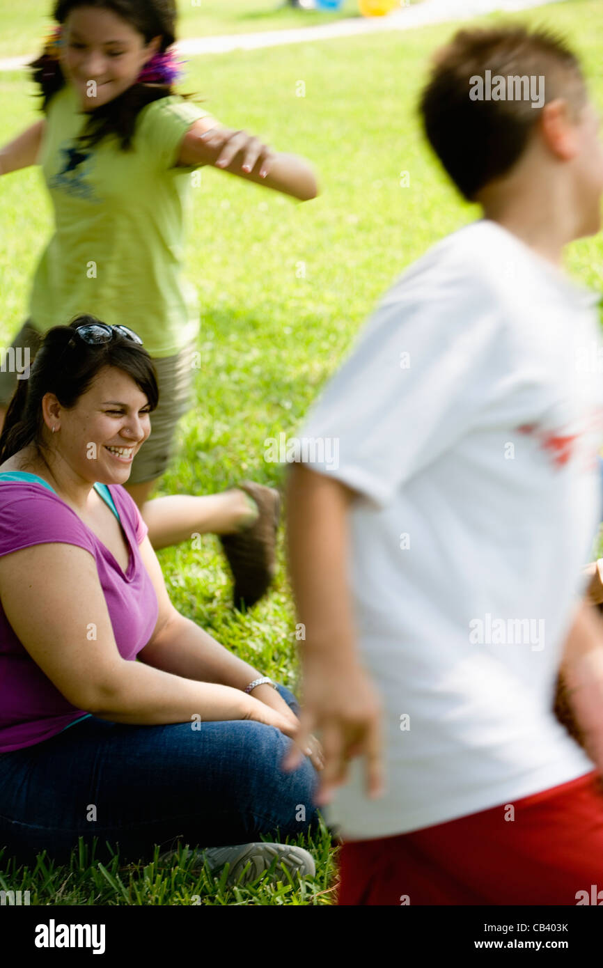 A boy and a girl running around their teacher sitting on the grass at a ...