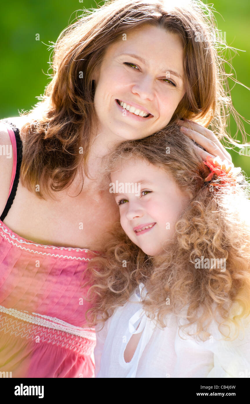 Beautiful and happy young mother hugging her daughter, both smiling, summer park in background ...