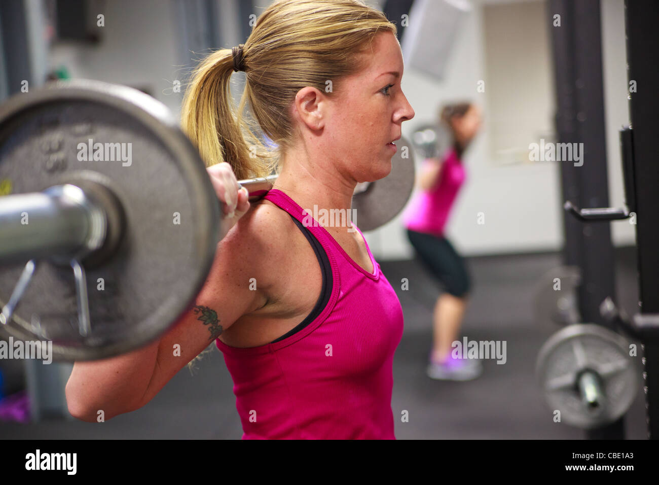 Blond women in pink top doing back squats with barbell in weight gym ...