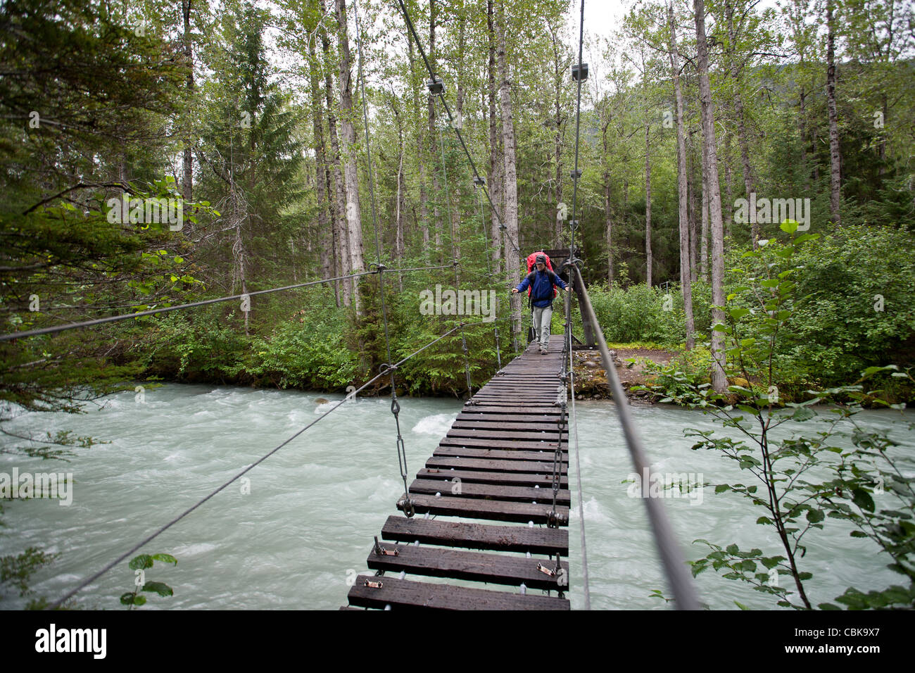 Backpacker crossing a suspension footbridge over Taiya river. Canyon ...