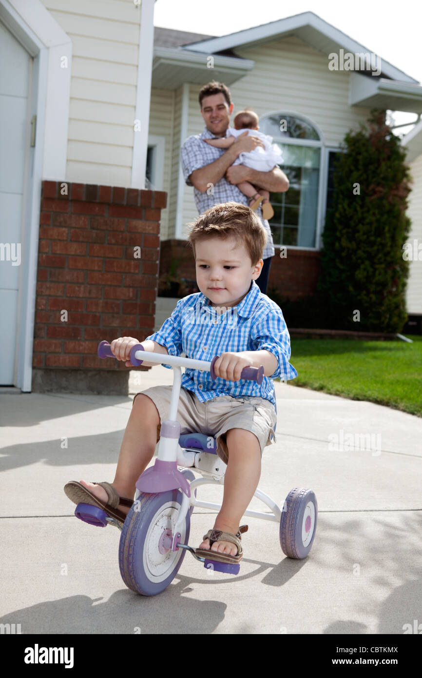 Toddler boy learning to ride tricycle with father in background Stock ...