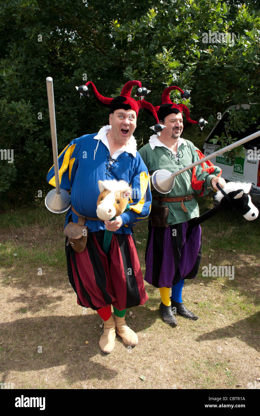 Two men in jester costume on hobby horses with wooden lances laughing ...