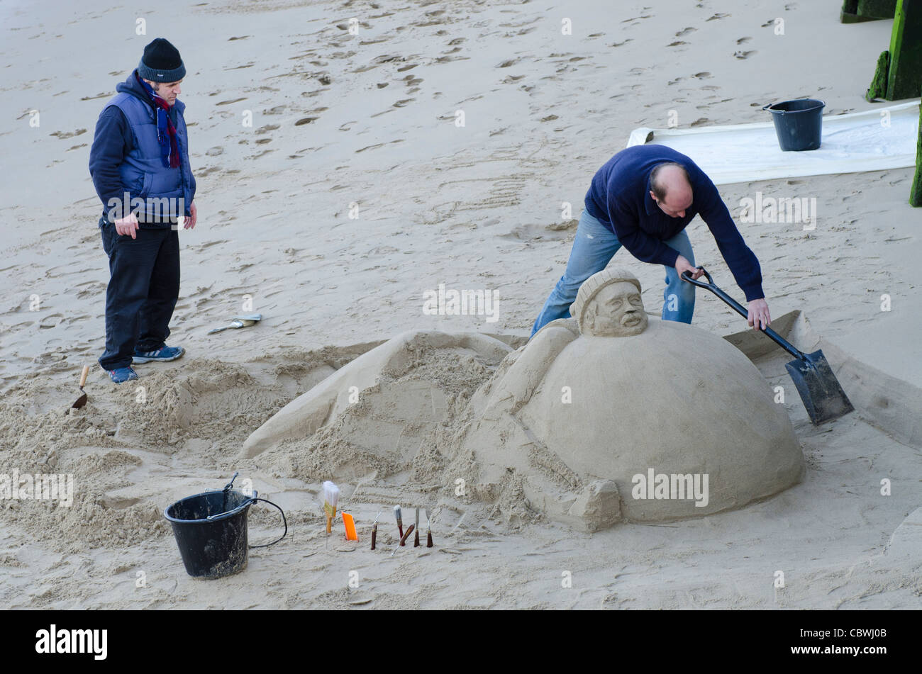 Two men working on Sand sculpture of head Shore South bank of river ...