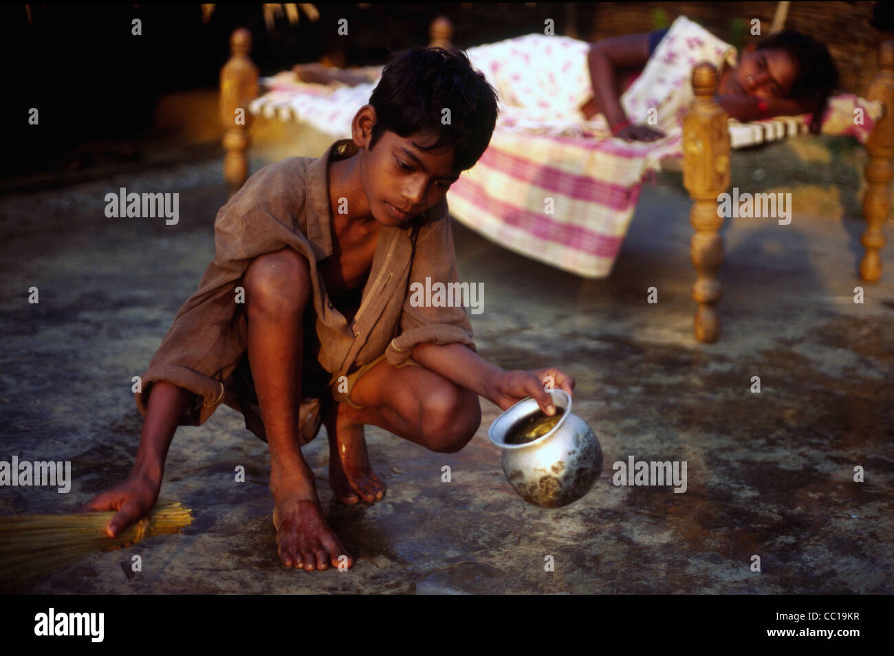 A boy washes and sweeps the floor while his mother lies on a charpoy ...