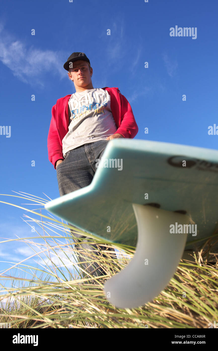 A surfer with hands in pockets stands over his surfboard against a blue ...