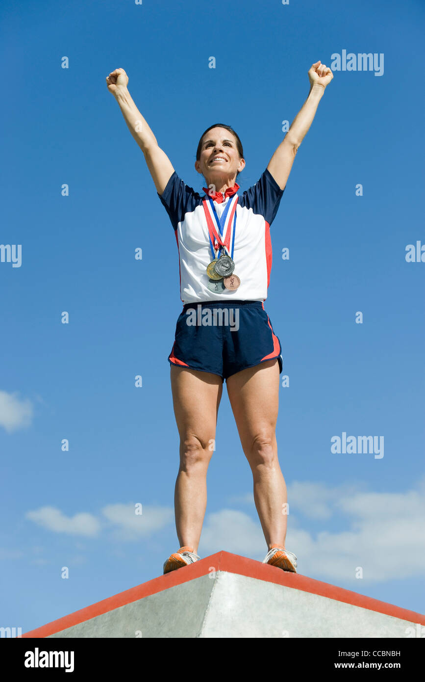 Female athlete standing on winner's podium with arms raised in victory ...
