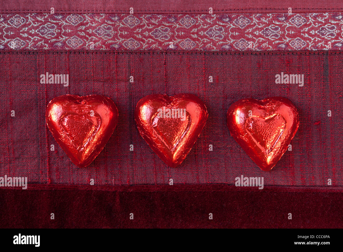 Three red foil wrapped candy hearts in a row on red textured background ...
