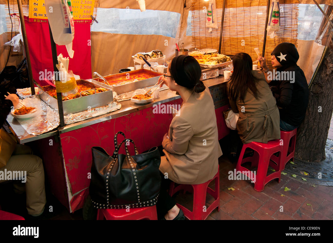 young women sitting down and eating from a street food stall near Ewha ...