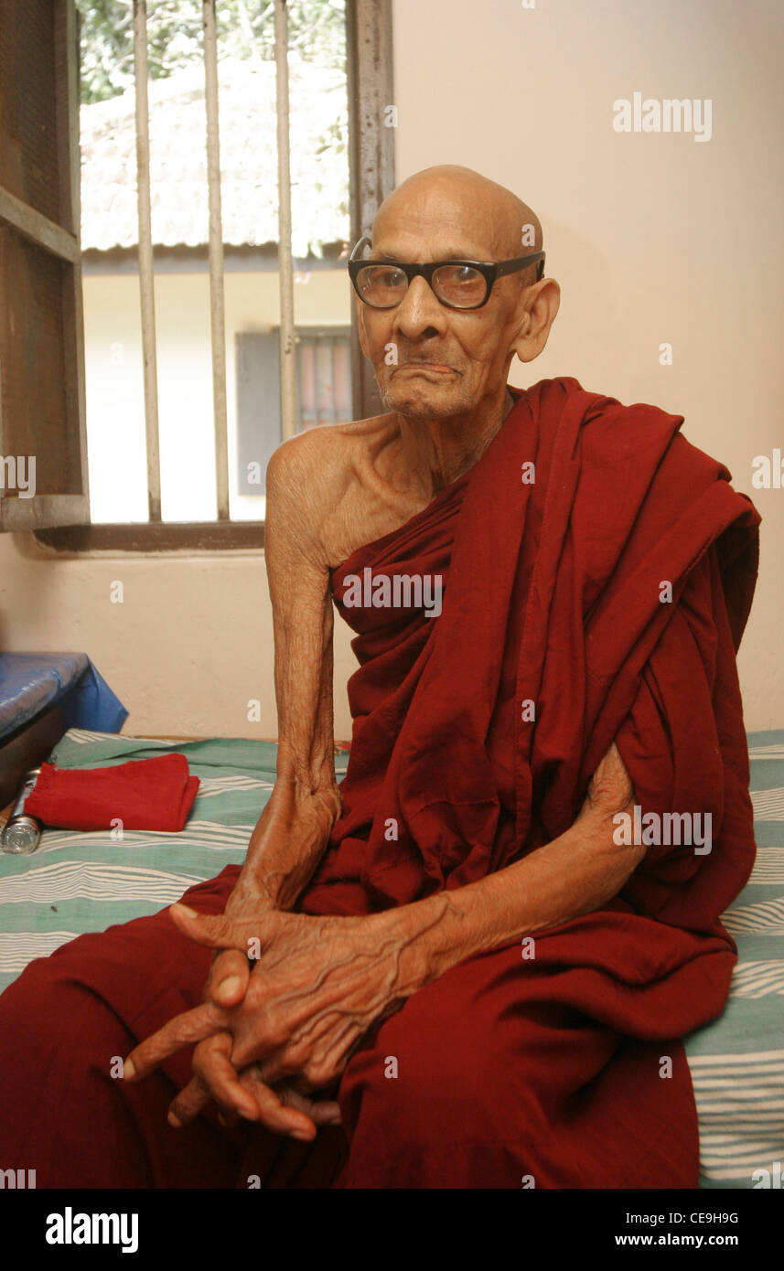 A elderly Buddhist Monk sits on the side of his bed in his room at a ...