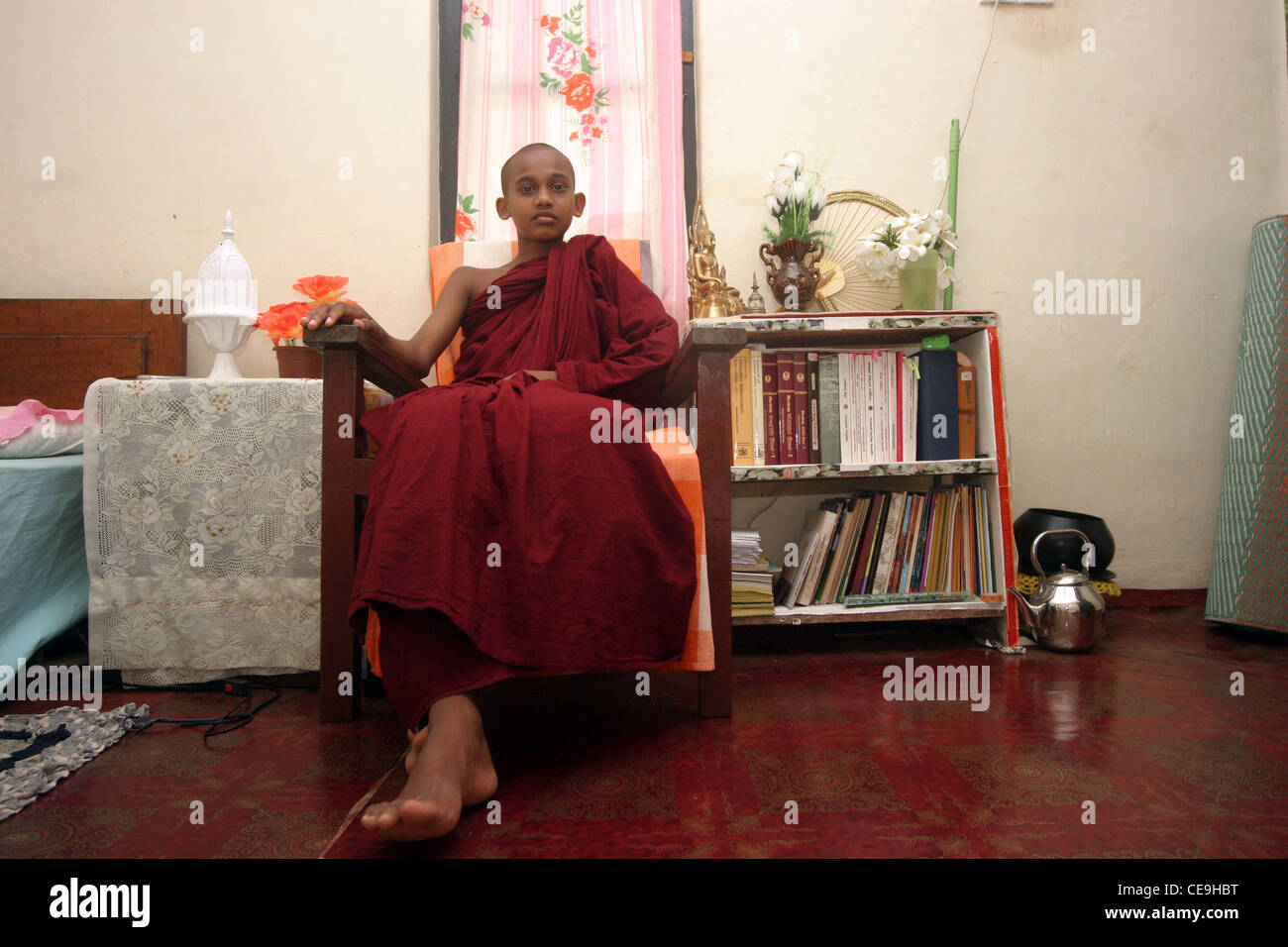 A young Buddhist Monk dressed in traditional red robes sits in his room ...