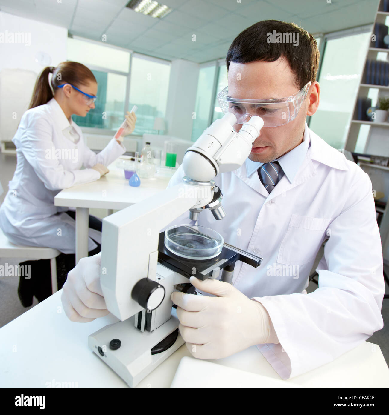 Male researcher looking through the microscope, his colleague in the ...