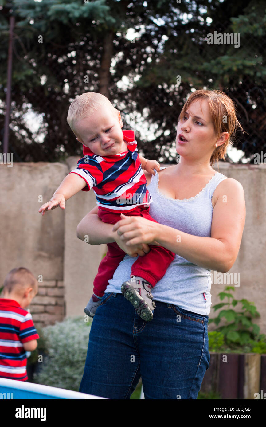 Young mother holding grumpy crying toddler boy outdoors in the garden ...