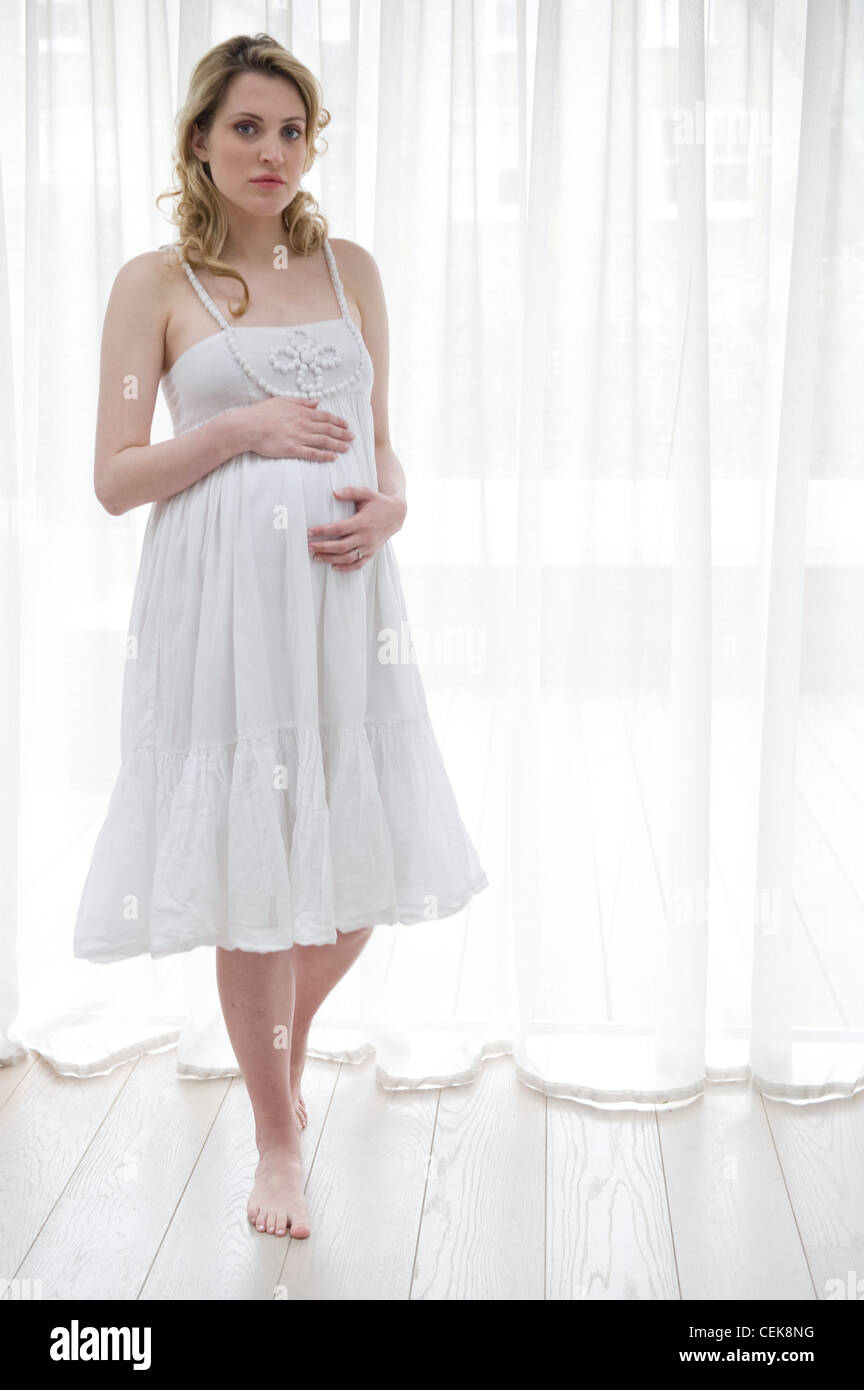 Pregnant Female Wavy Blonde Hair Wearing A Knee Length White Summer Dress Standing By The