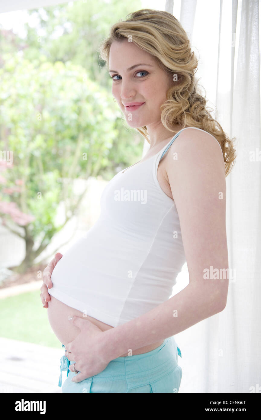Pregnant Female Wavy Blonde Hair Wearing A White Vest And Blue Trousers Standing By The Window