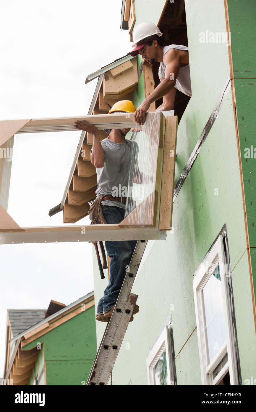 Hispanic carpenter on ladder handing window frame to second carpenter ...