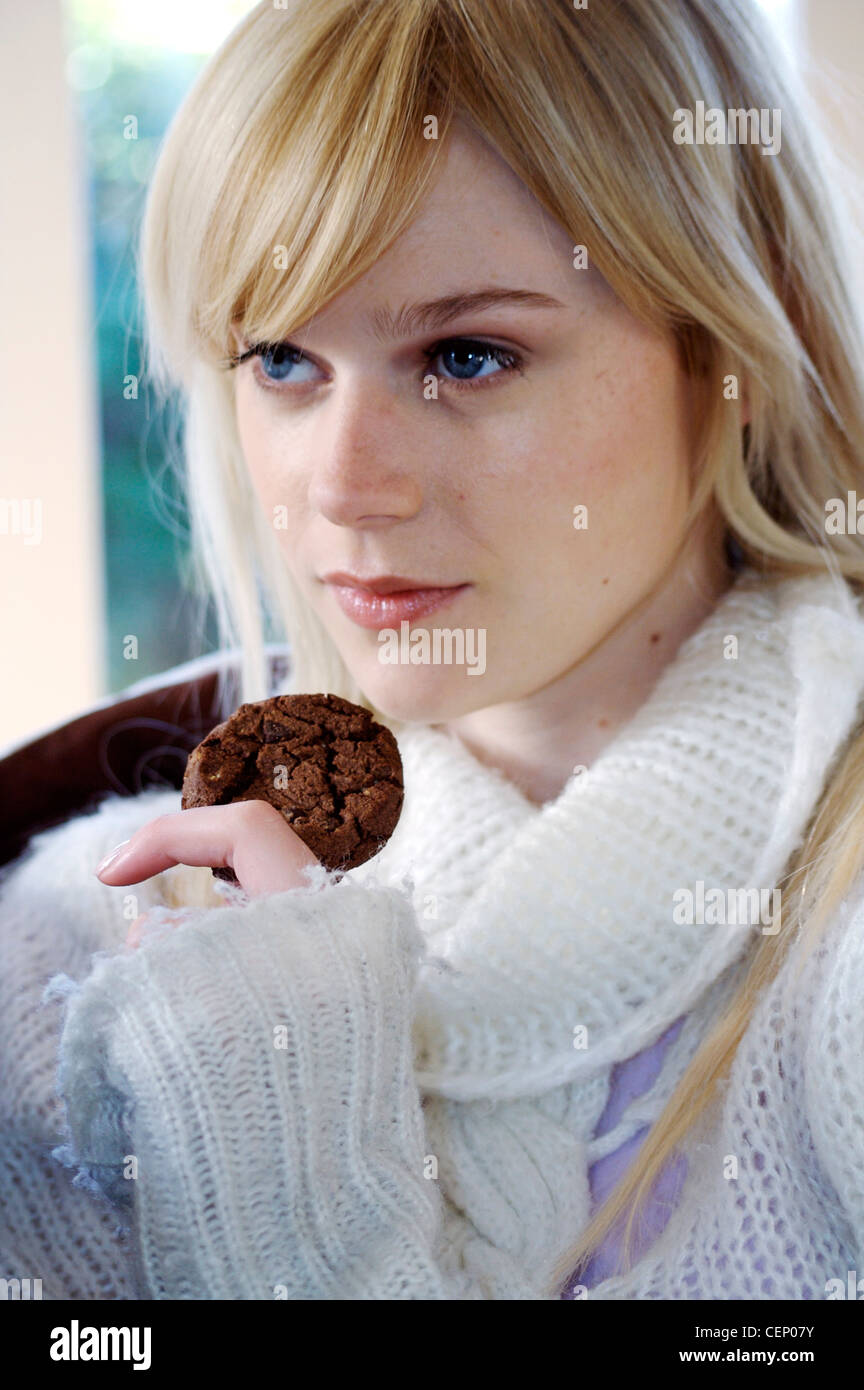 Female wearing white knitted jumper, eating chocolate cookie, looking ...