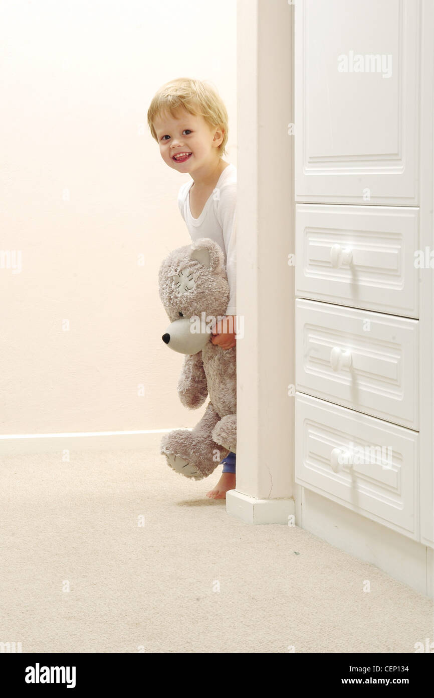 Blonde boy peering around wall down hallway, holding a grey teddy bear ...