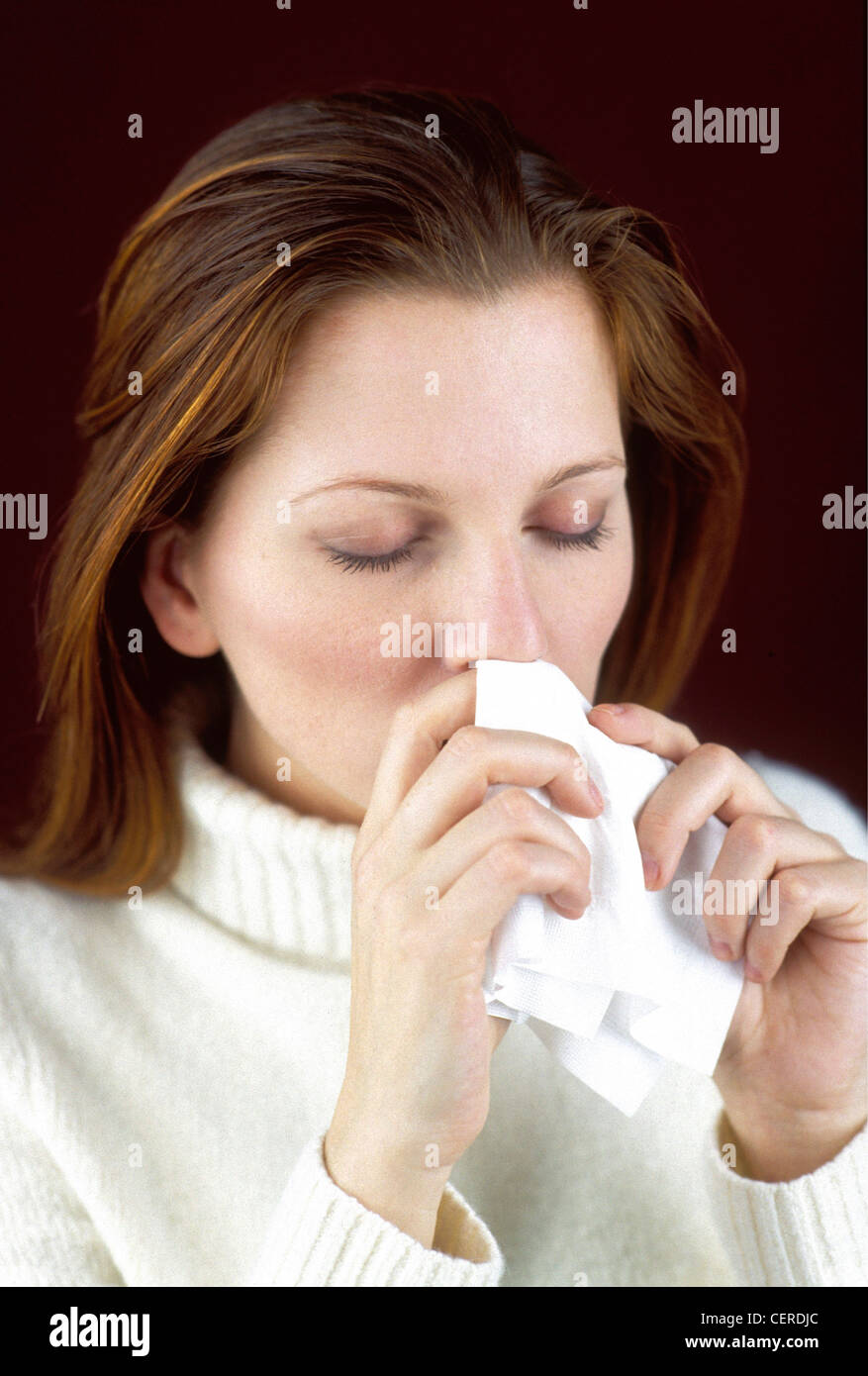 Brunette female wiping nose with a tissue, wearing a cream polo neck ...