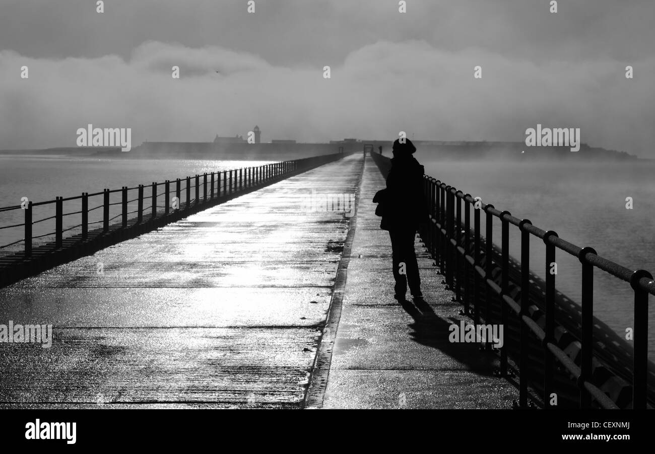 A lone silhouetted figure walking towards a lighthouse along a wet ...