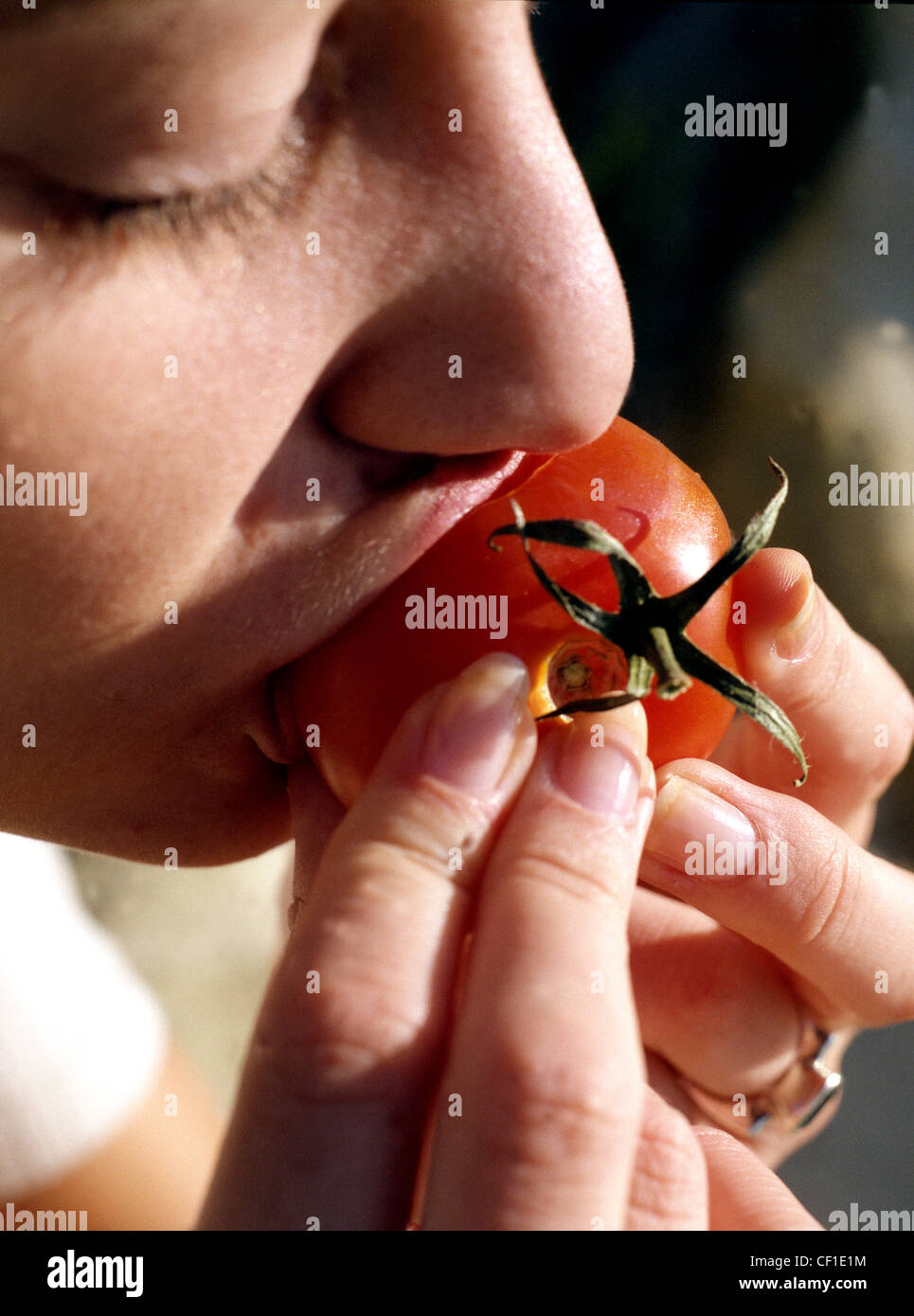A female holding a red tomato, biting into it, with some large rocks in ...