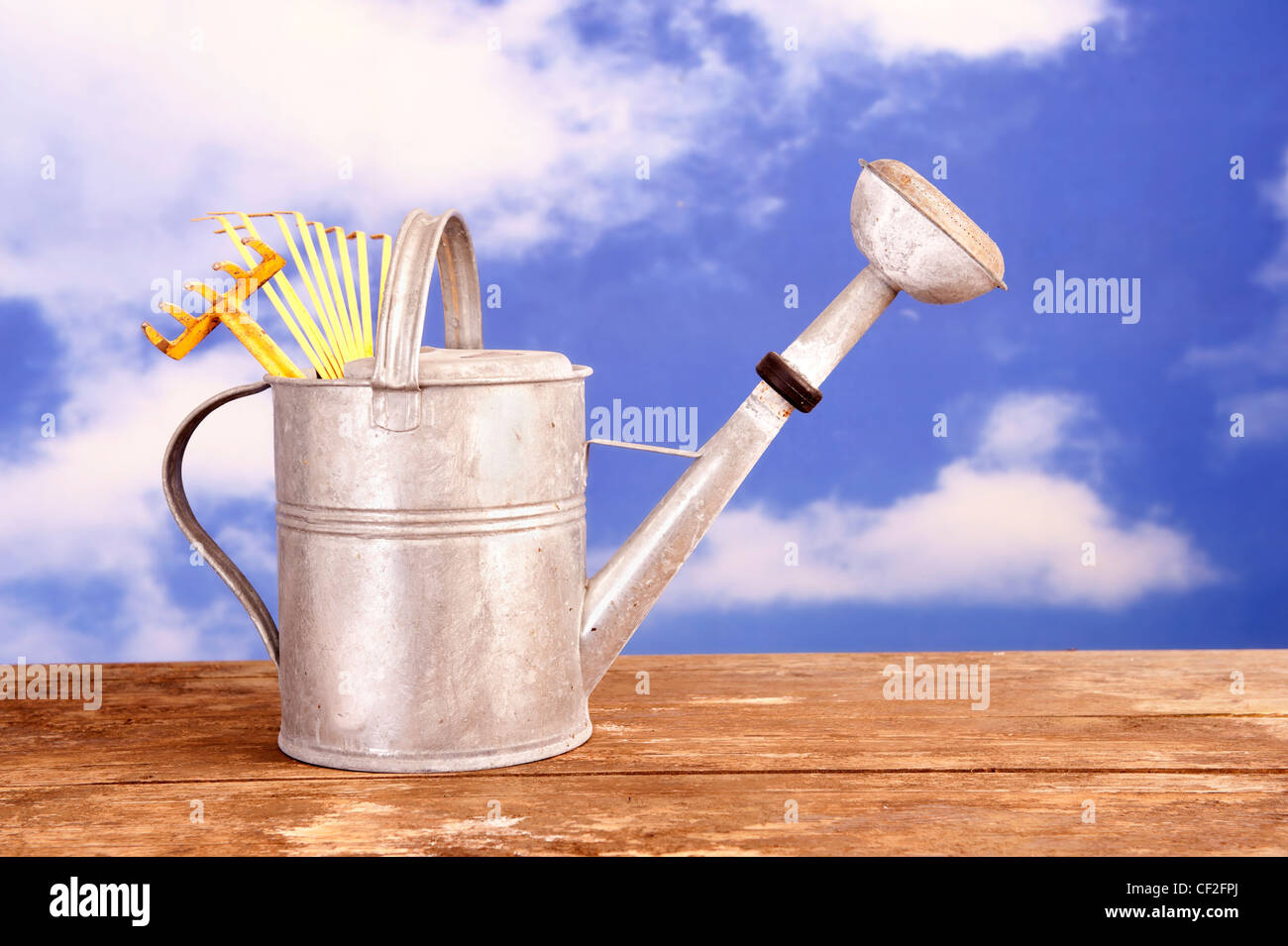 metal watering can with yellow rakes against a blue sky with clouds ...