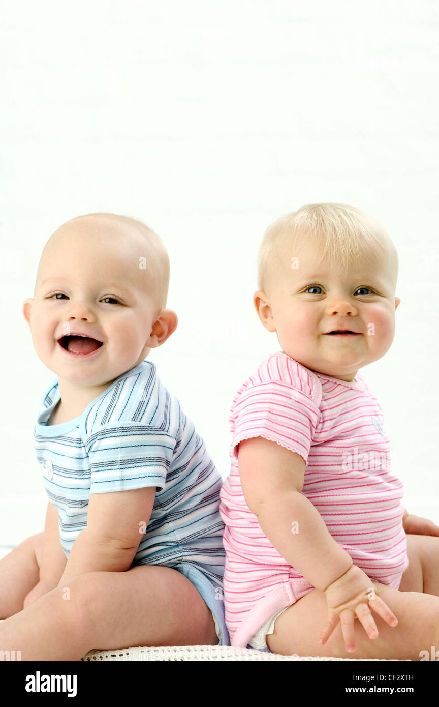 Two babies sitting back to back, male baby on left wearing blue striped ...