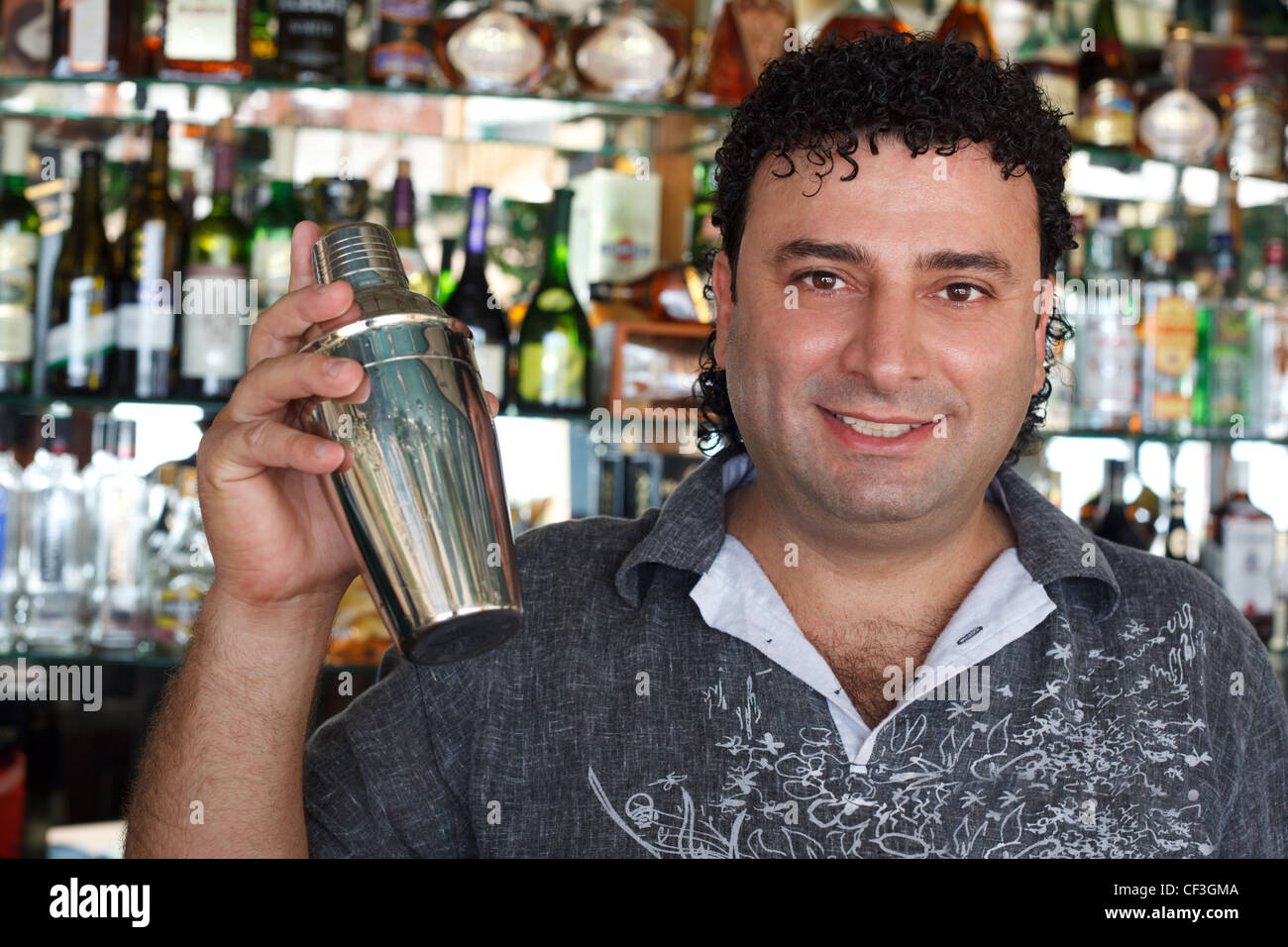 Barman with shaker behind bar rack. Smiling man against shelves with ...