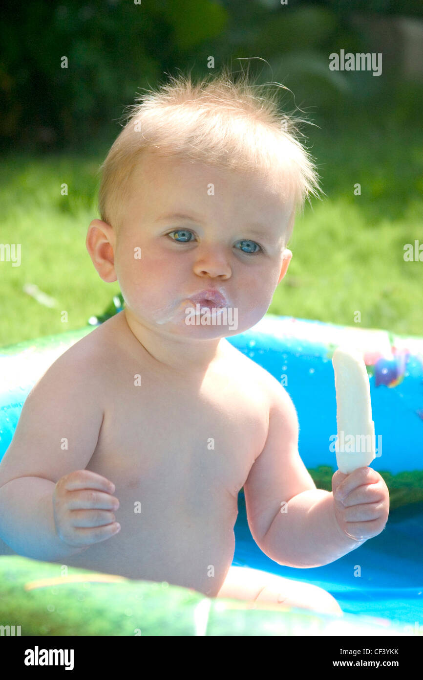 Baby sitting in paddling pool eating milk pop ice lolly, looking to ...