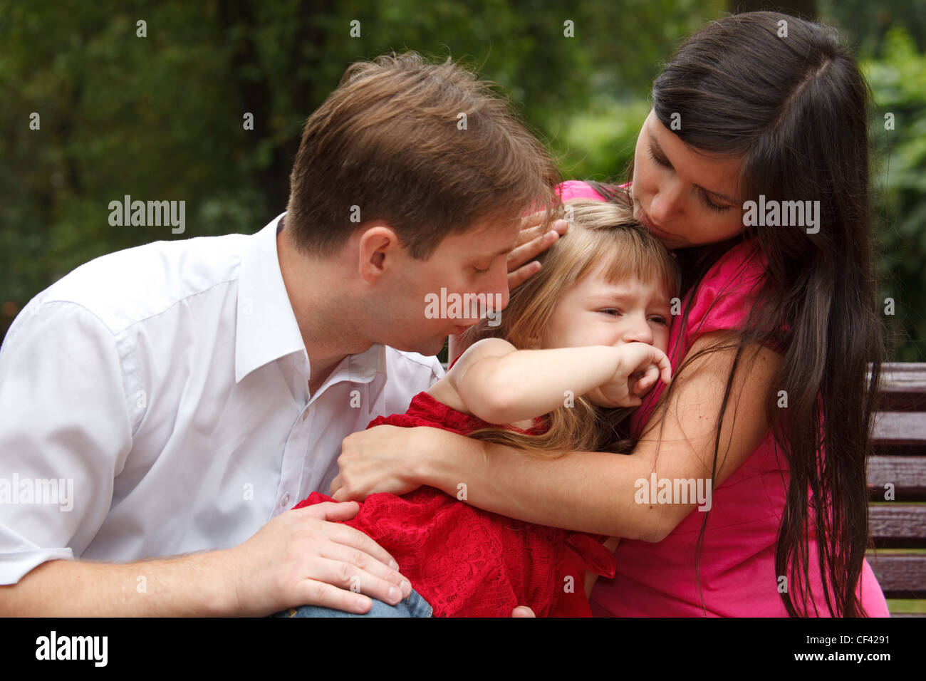 Parents calm crying girl on walk in summer garden. Mum embraces ...