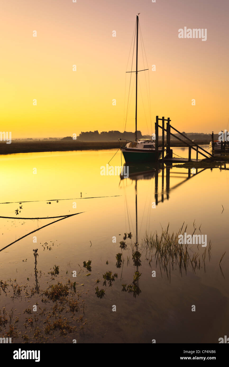 A boat moored alongside a jetty, reflected in still water at sunrise ...