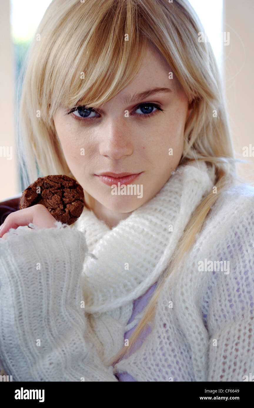Female wearing white knitted jumper, eating chocolate cookie, looking ...