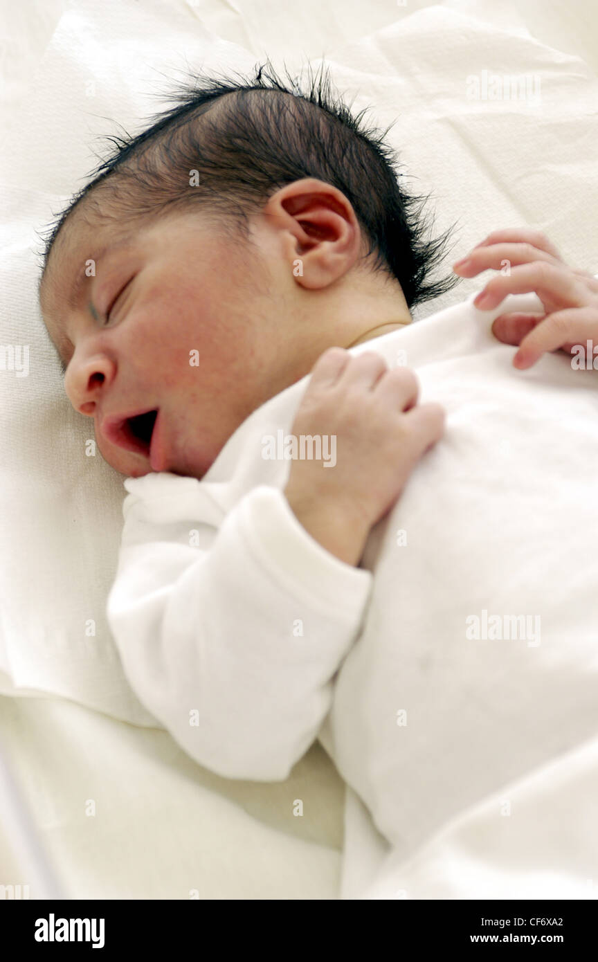 Newborn baby wearing white babygrow lying on back hands up head turned ...