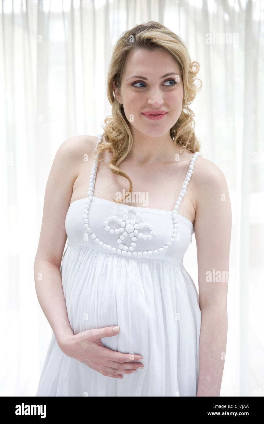 Pregnant Female Wavy Blonde Hair Wearing A White Dress Standing By The Window Hand On Bump