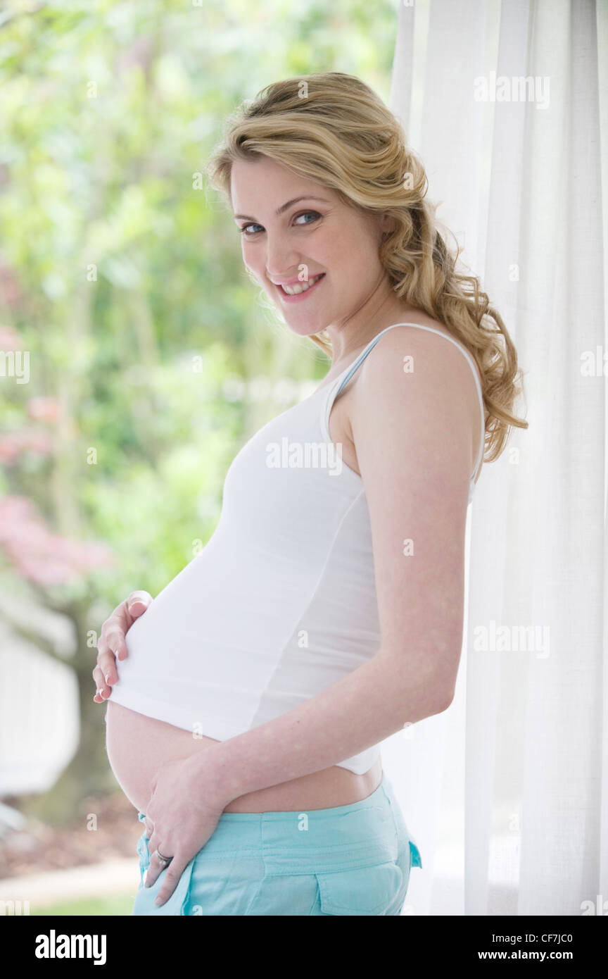 Pregnant Female Wavy Blonde Hair Wearing A White Vest And Blue Trousers Standing By The Window